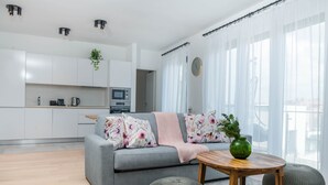 Uma sala de estar moderna com um sofá cinzento, uma mesa de centro de madeira e pufes cinzentos. Uma cozinha com armários brancos e um micro-ondas é visível ao fundo.