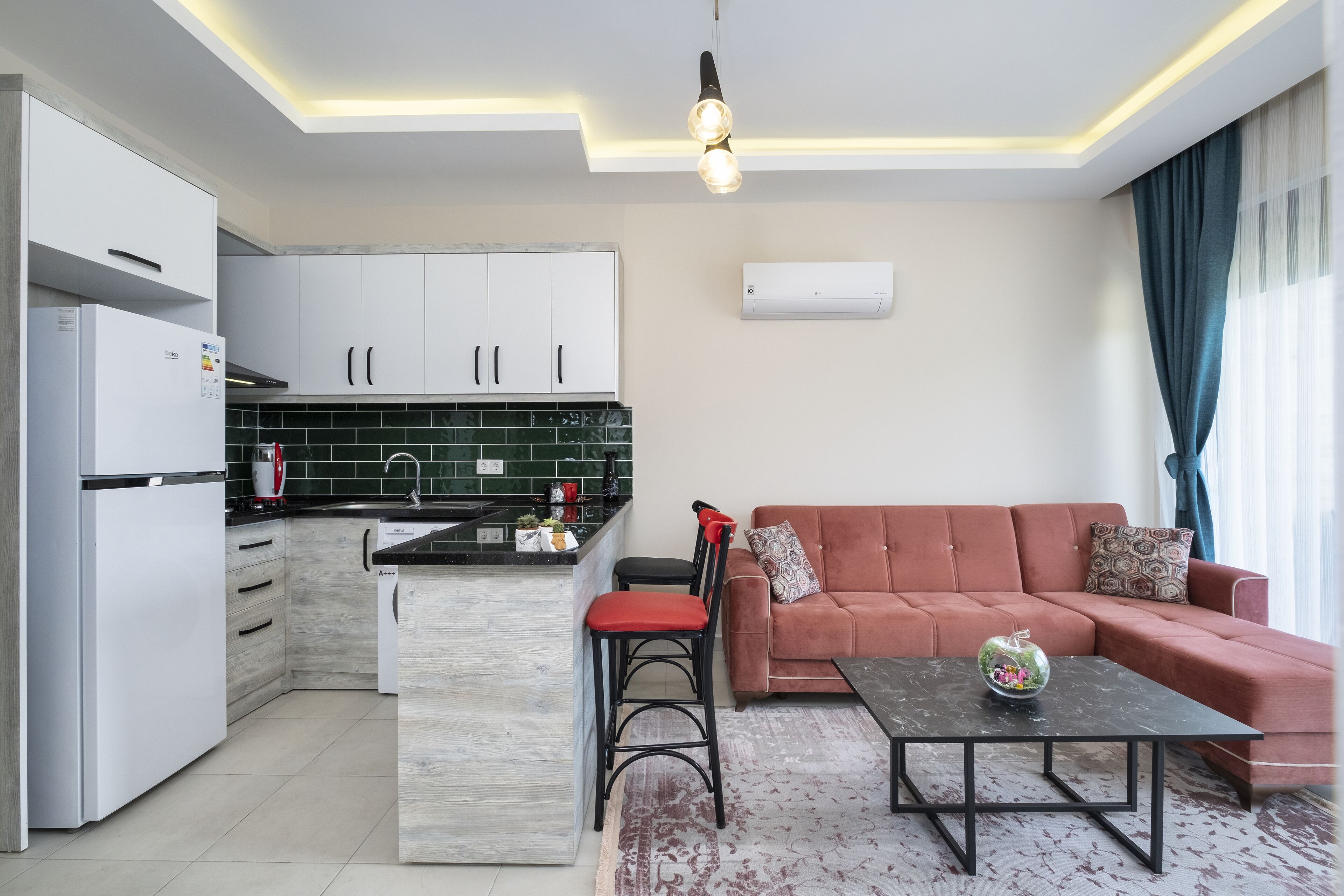 Uma cozinha moderna com armários brancos, bancada preta e uma cadeira vermelha. Há um sofá com almofadas estampadas e uma mesa de centro com um objeto decorativo.
