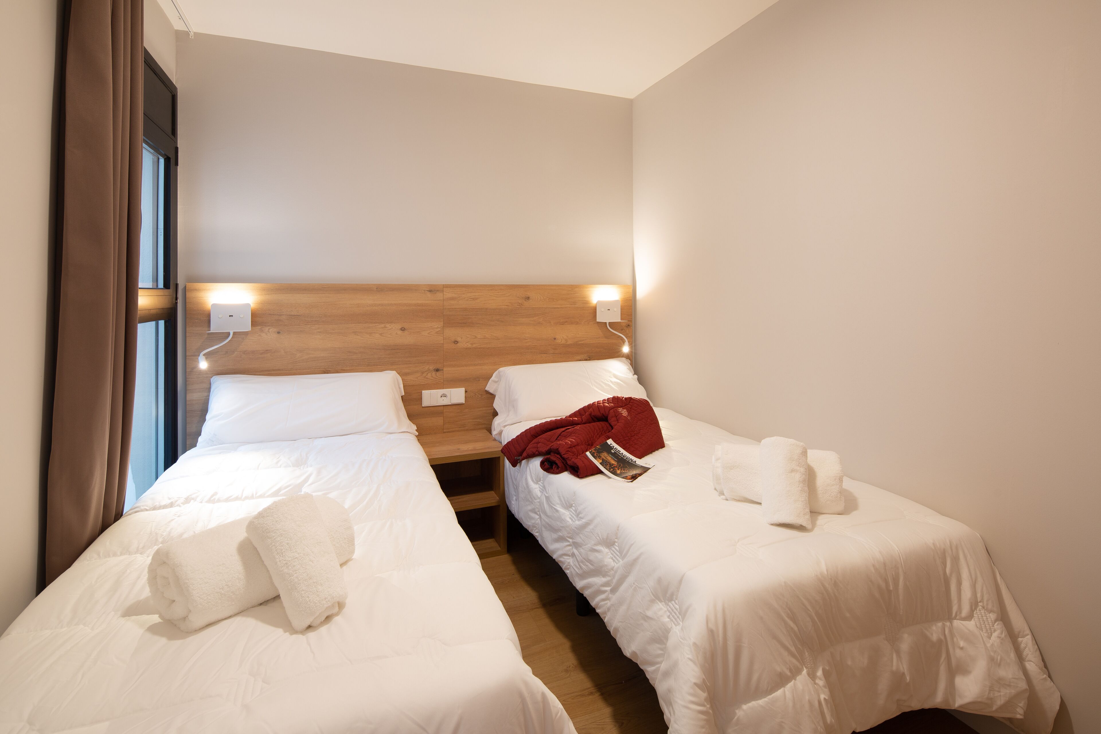 Quarto de hotel com duas camas de solteiro, cabeceiras de madeira, roupa de cama branca e uma mesinha de cabeceira com abajur.