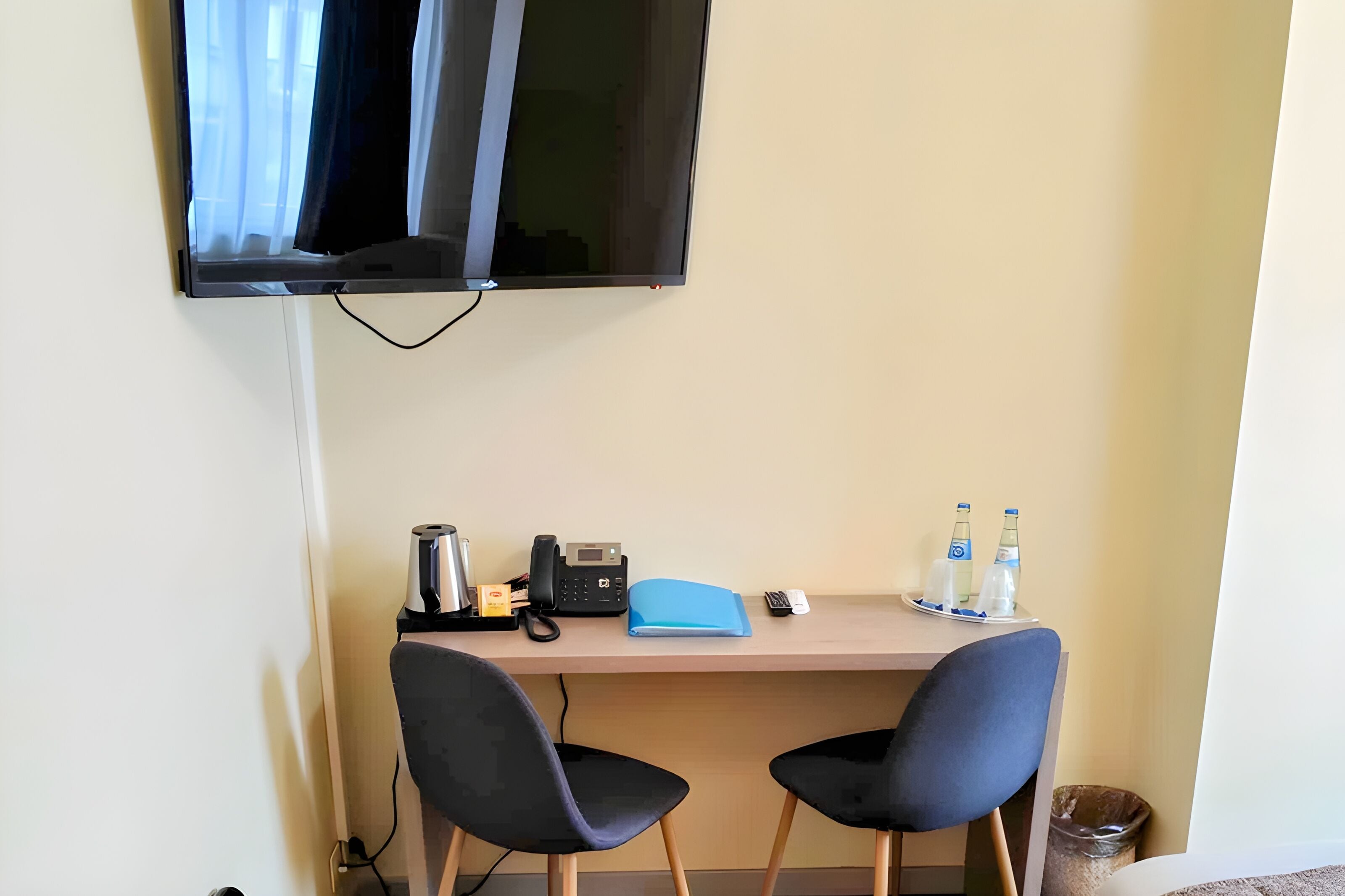 Um pequeno espaço de trabalho com uma mesa, duas cadeiras, uma televisão montada na parede, uma máquina de café, um telefone, uma pasta azul e duas garrafas de água.