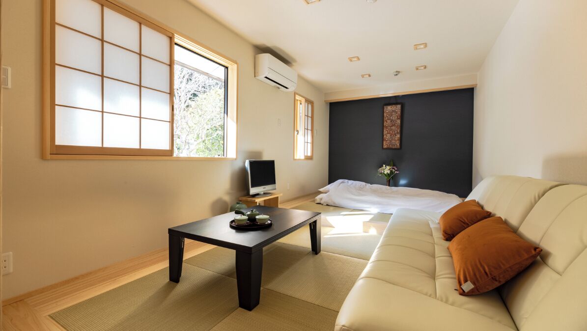 Quarto com piso de tatame, cama com roupa de cama branca, sofá com almofadas laranja, televisão e uma pequena mesa com um objeto decorativo.