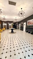 Um hall com piso de padrão hexagonal, uma mesa redonda central, cadeiras amarelas e várias luminárias.