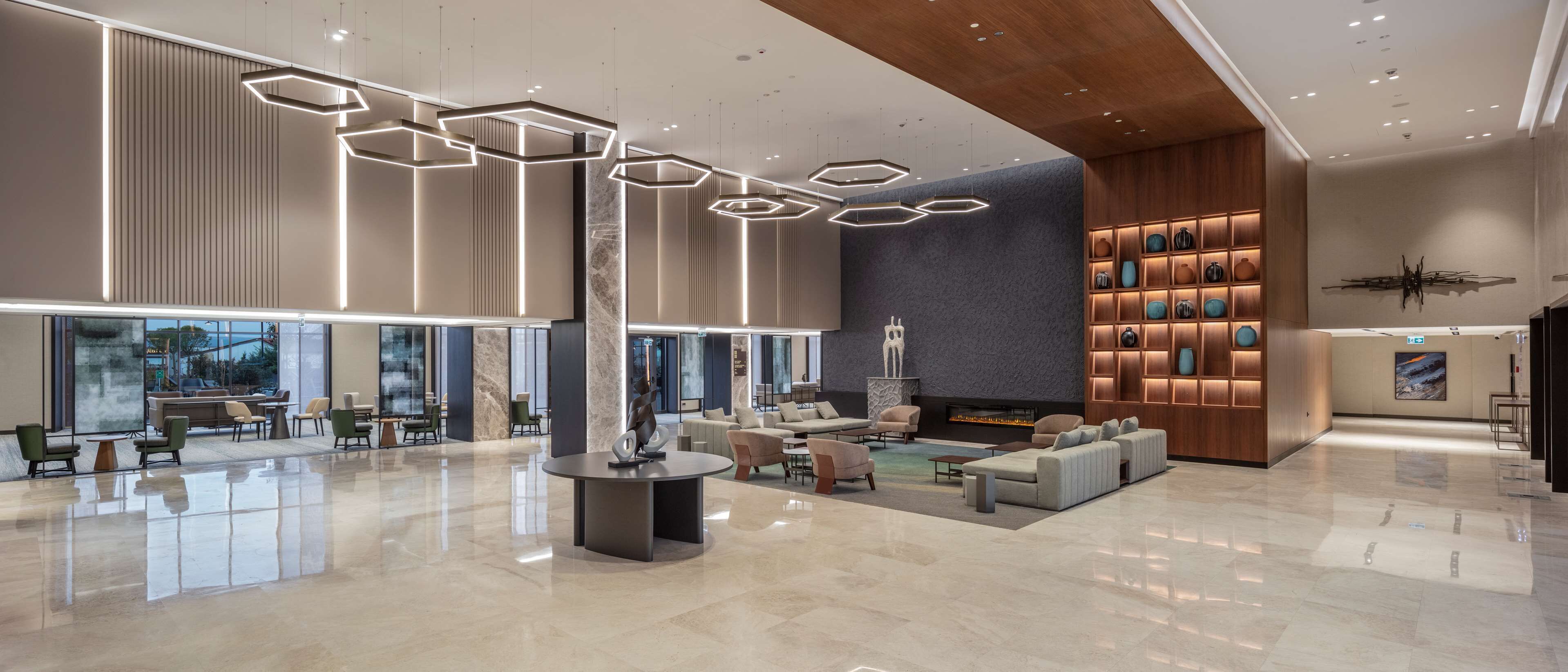 Um hall amplo com teto alto, luminárias modernas, áreas de estar e um painel decorativo na parede.