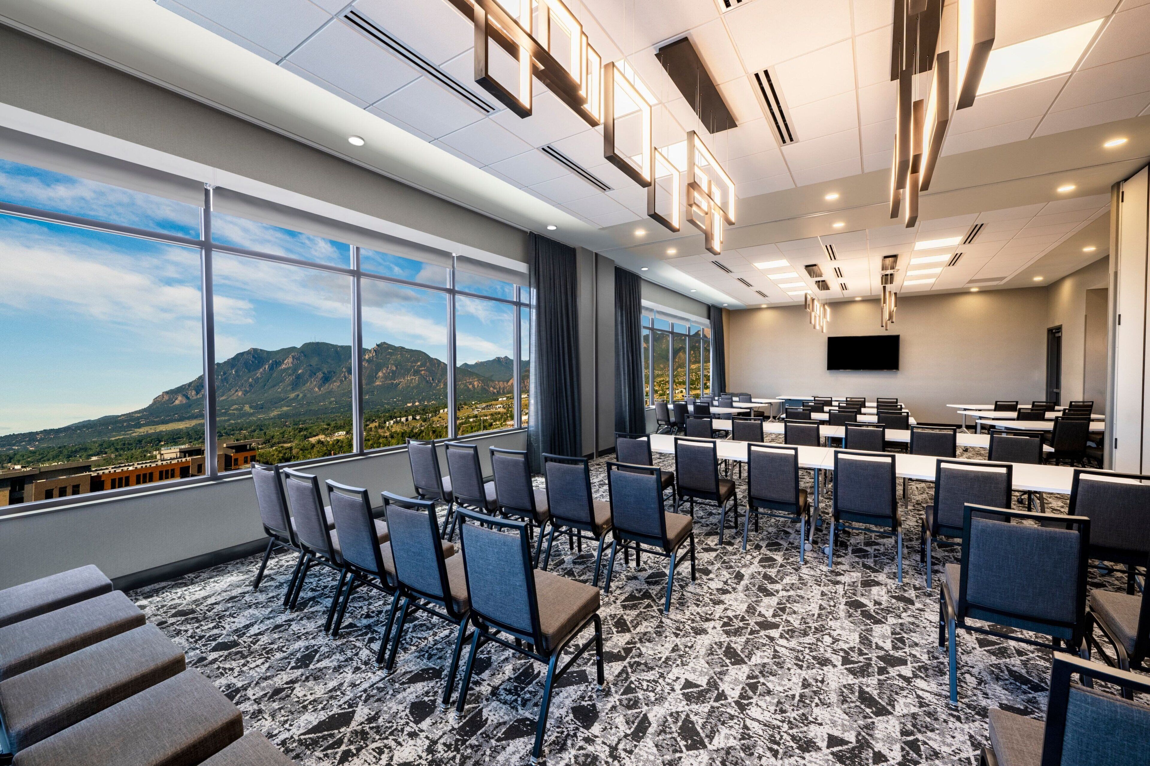 Uma sala de conferências com vista panorâmica das montanhas, equipada com fileiras de cadeiras e mesas, e uma TV de tela plana na parede.