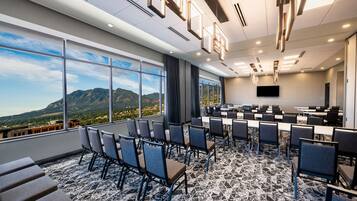 Uma sala de conferências com vista panorâmica das montanhas, equipada com fileiras de cadeiras e mesas, e uma TV de tela plana na parede.