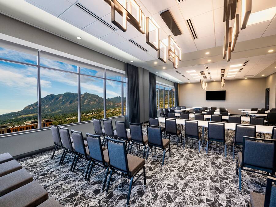 Uma sala de conferências com vista panorâmica das montanhas, equipada com fileiras de cadeiras e mesas, e uma TV de tela plana na parede.