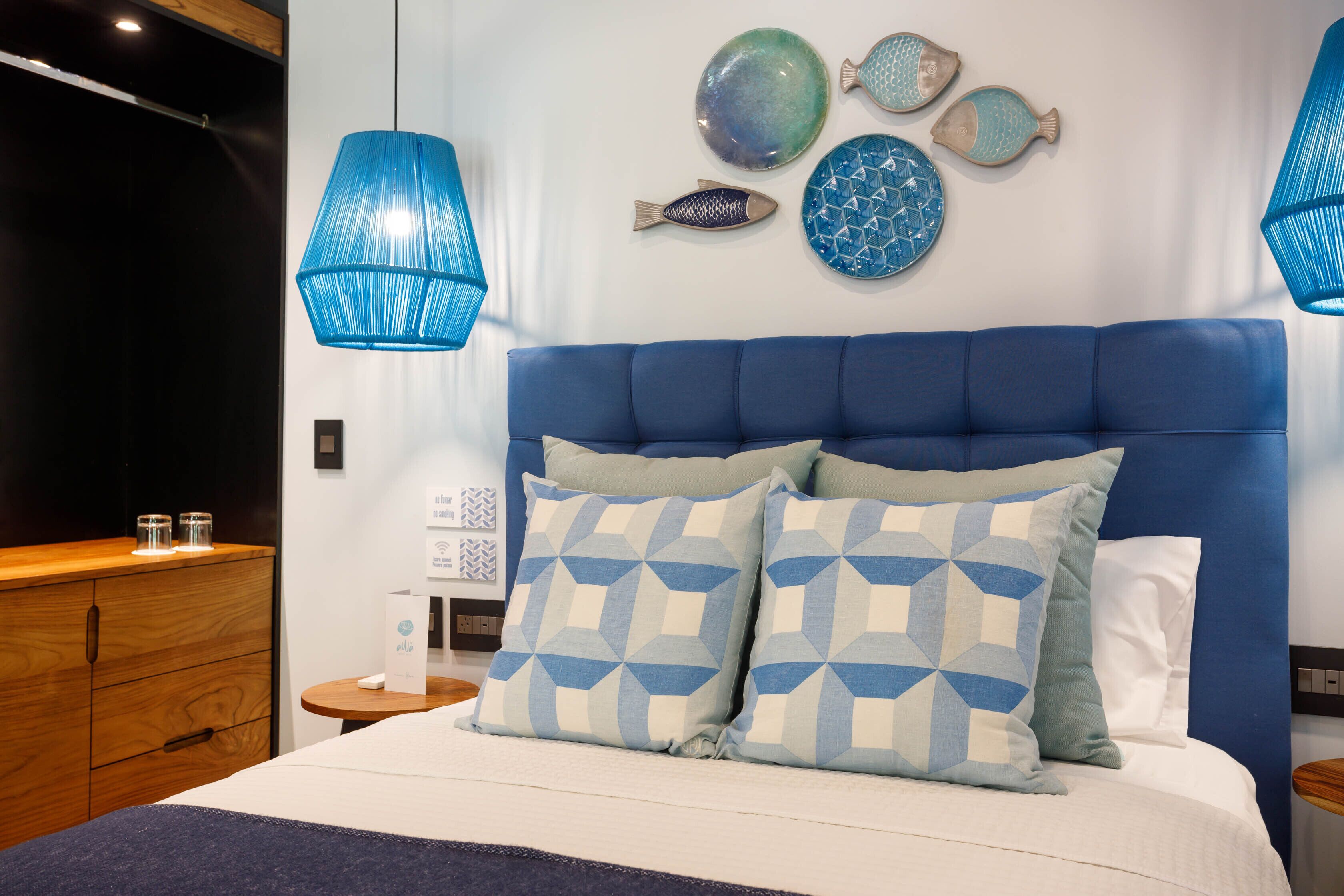 Quarto de hotel bem arrumado com cabeceira estofada azul, almofadas estampadas com padrão geométrico e pratos decorativos de peixe na parede.