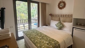 Quarto de hotel com cama, escrivaninha com cadeira, ar condicionado, pequeno frigorífico e varanda com vista para o verde.