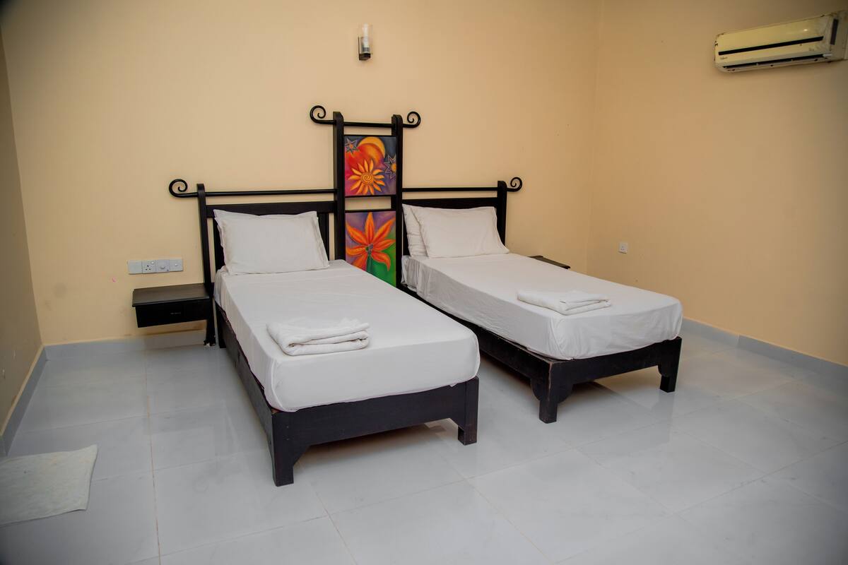 Quarto com duas camas de solteiro, mesa de cabeceira, ar condicionado fixado na parede e um quadro decorativo.