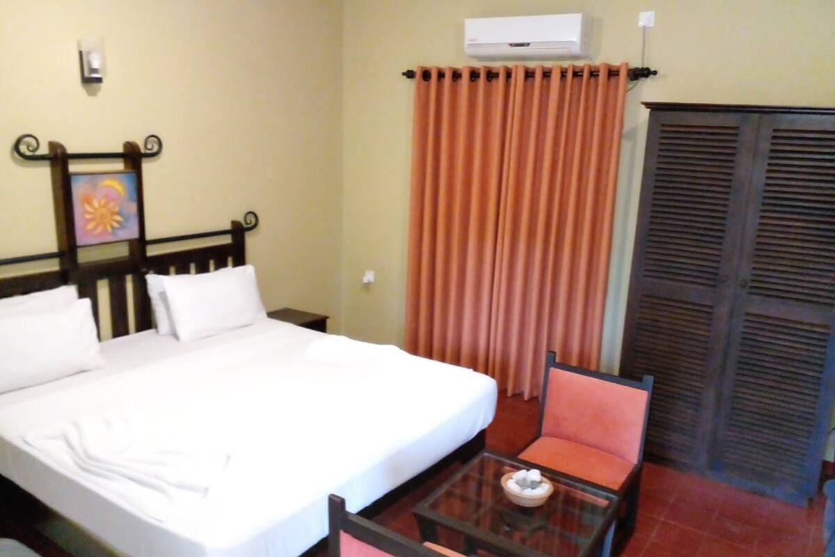 Quarto de hotel com cama, cadeira, mesinha, ar condicionado e janela com cortinas.