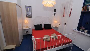 Quarto com colcha vermelha, cabeceira branca, mesas de cabeceira com abajures, um guarda-roupa e um banquinho pequeno.