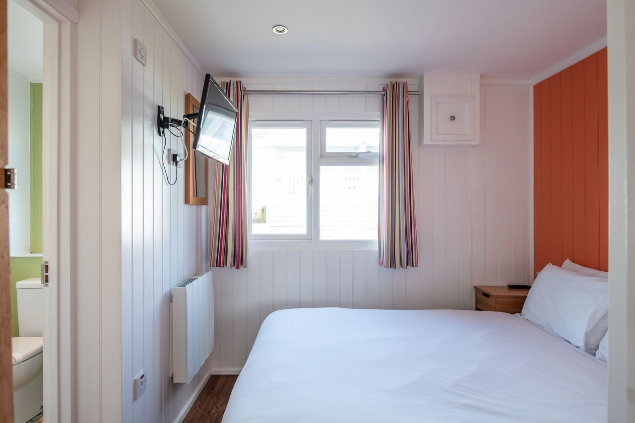 Quarto com cama, televisão na parede, janela com cortinas, radiador e mesa de cabeceira.