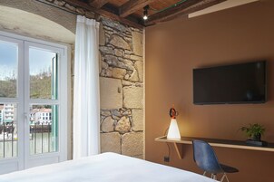 Quarto com parede de pedra, TV fixada na parede, mesa de cabeceira com abajur, uma cadeira e janela com vista para edifícios e água.