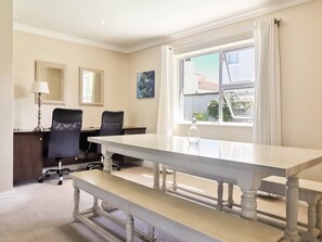 Uma sala de conferências com uma mesa comprida, duas cadeiras, uma escrivaninha com abajur e uma janela com vista para edifícios e vegetação.