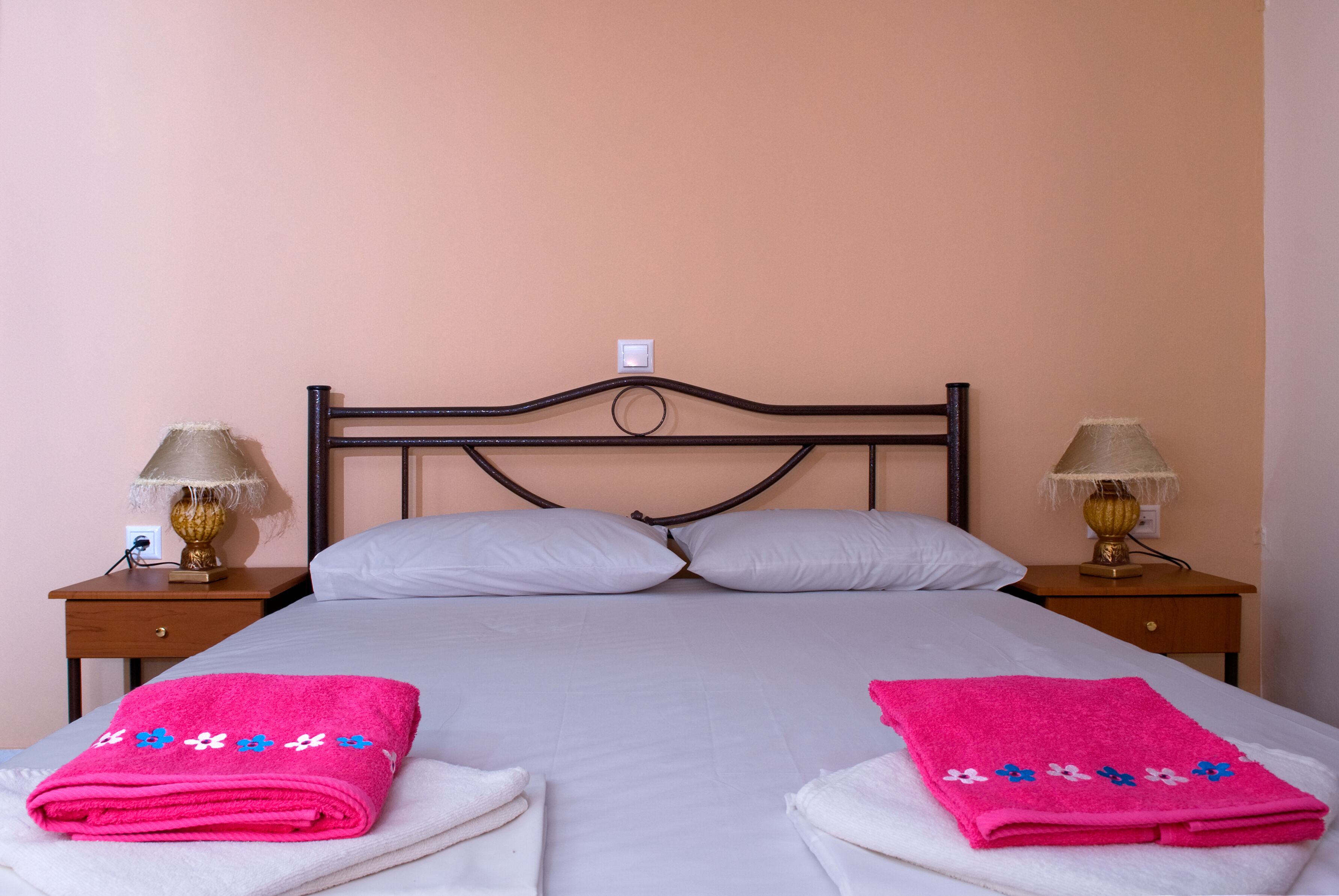 Cama bem arrumada com duas toalhas cor-de-rosa, cabeceira de madeira e duas luminárias em mesas de cabeceira.