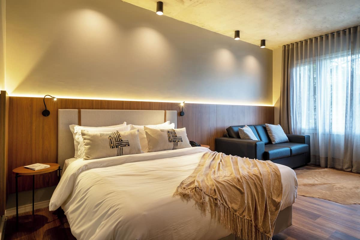 Quarto de hotel com uma cama grande, um sofá, uma mesinha e uma janela com cortinas.