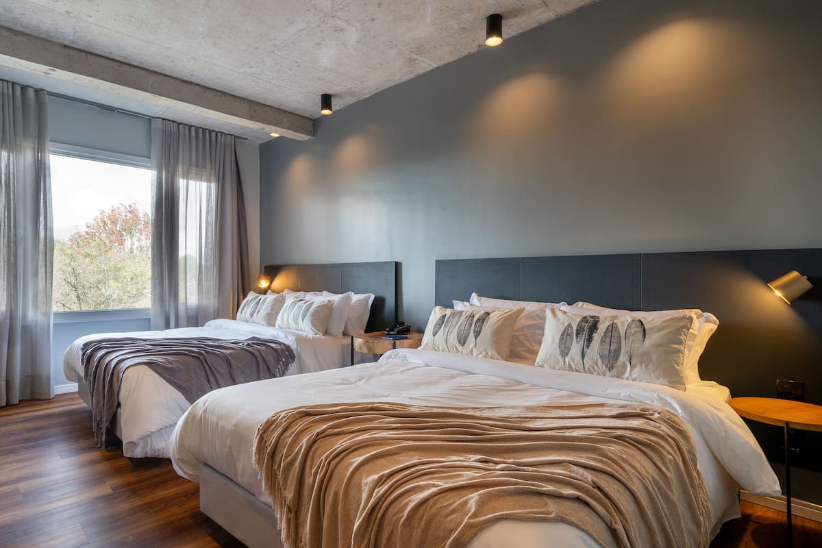 Quarto de hotel moderno com duas camas, uma janela grande com cortinas e vista para árvores.