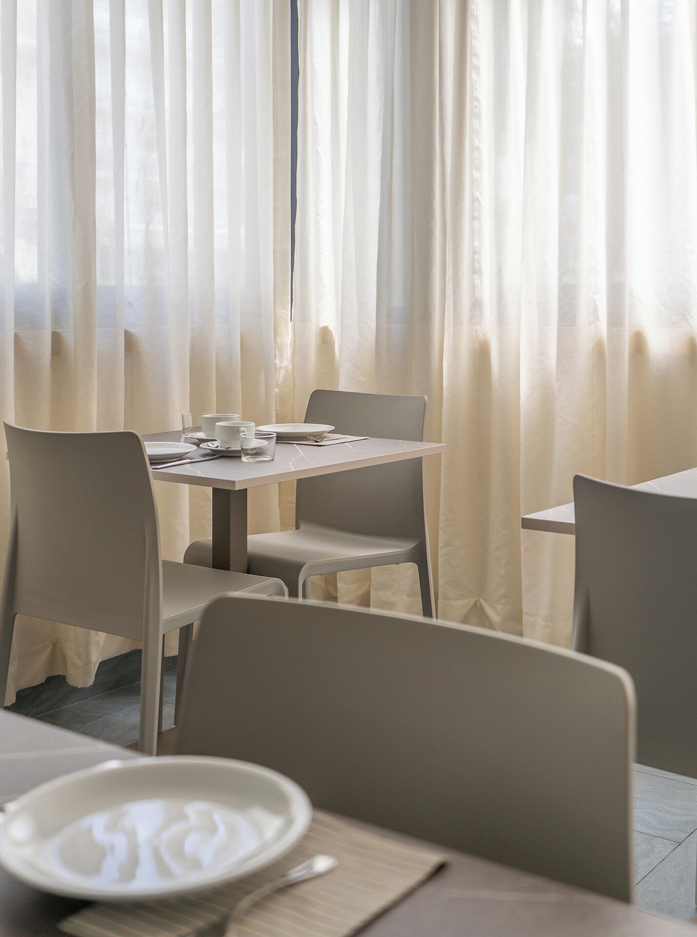 Uma área de refeições com uma mesa posta para dois, cadeiras e vista através de cortinas leves.