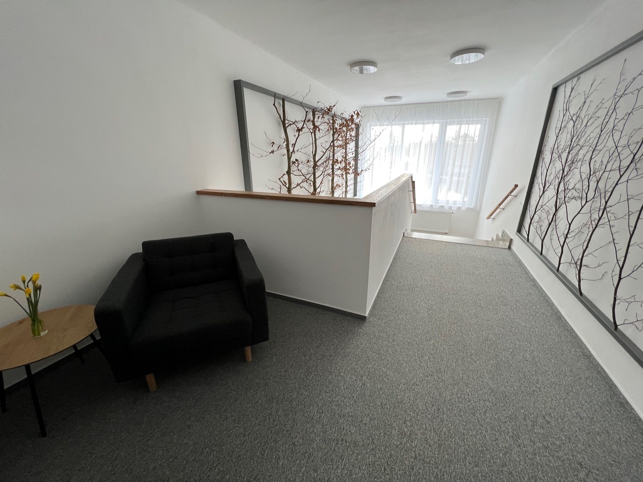 Um espaço de escritório moderno com uma mesa de recepção, uma cadeira de escritório preta, uma pequena mesa com um vaso e uma janela grande com cortinas leves.
