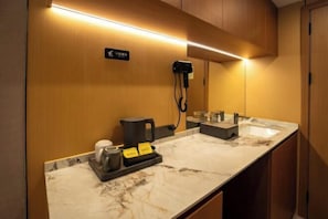 Casa de banho de hotel com bancada de mármore, máquina de café, secador de cabelo e porta-toalhas.