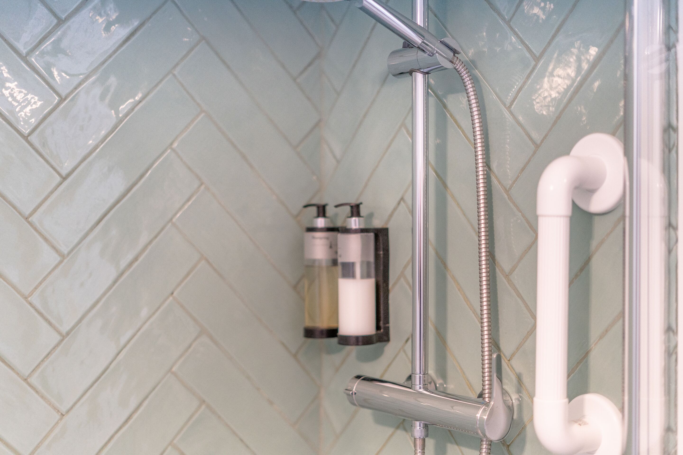 Chuveiro com torneira cromada e mangueira flexível, em frente a uma parede revestida com azulejos em padrão herringbone.