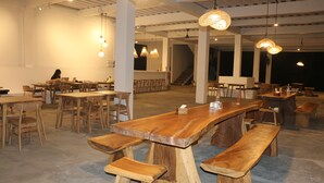 Um restaurante espaçoso com mesas e bancos de madeira, luminárias pendentes e uma escada visível ao fundo.