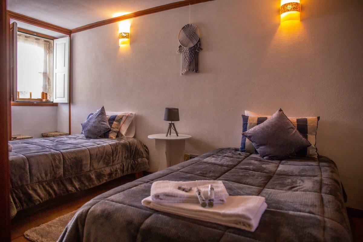 Quarto de hotel com duas camas, um ornamento circular na parede, uma mesinha com abajur e uma janela com cortinas.