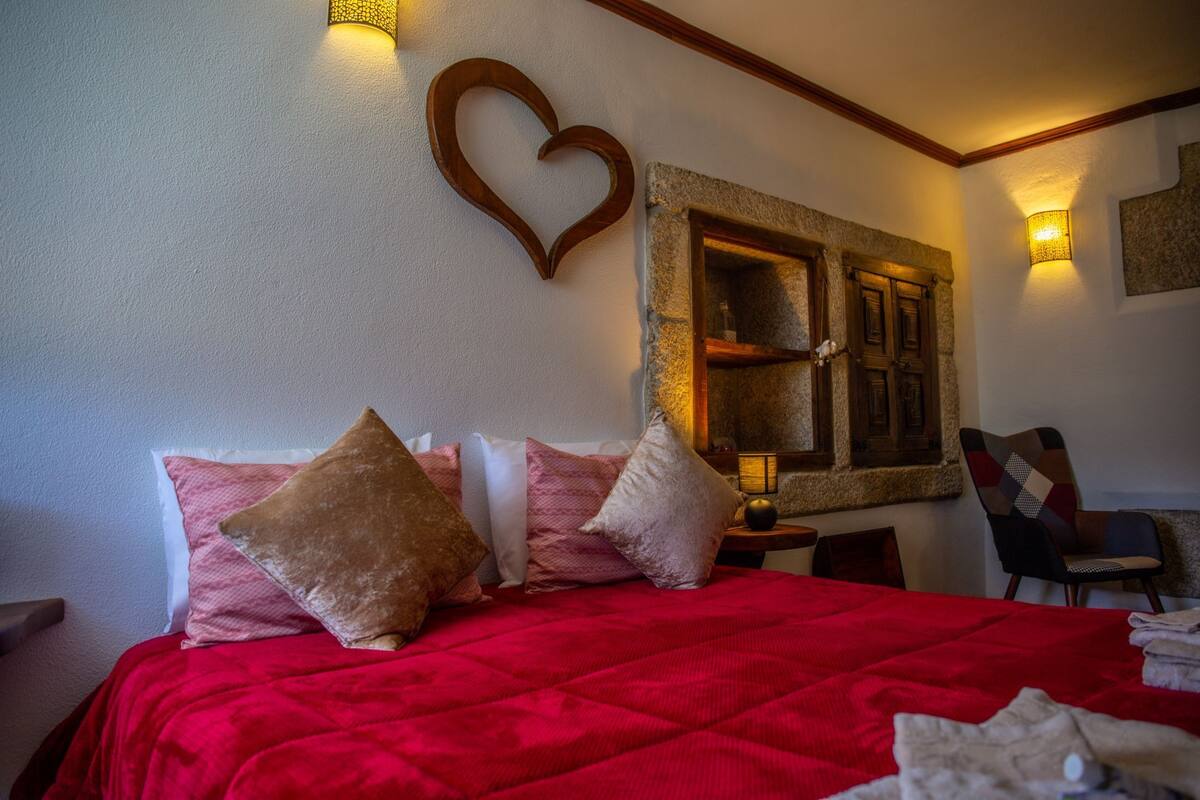 Quarto de hotel com uma cama grande, várias almofadas, uma decoração em formato de coração na parede, um espelho com moldura de pedra e uma cadeira.