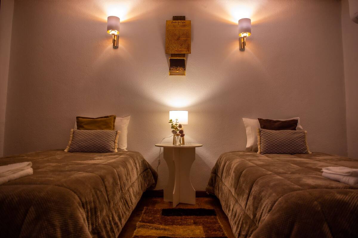 Quarto de hotel com duas camas, uma mesinha com abajur e luminárias na parede.