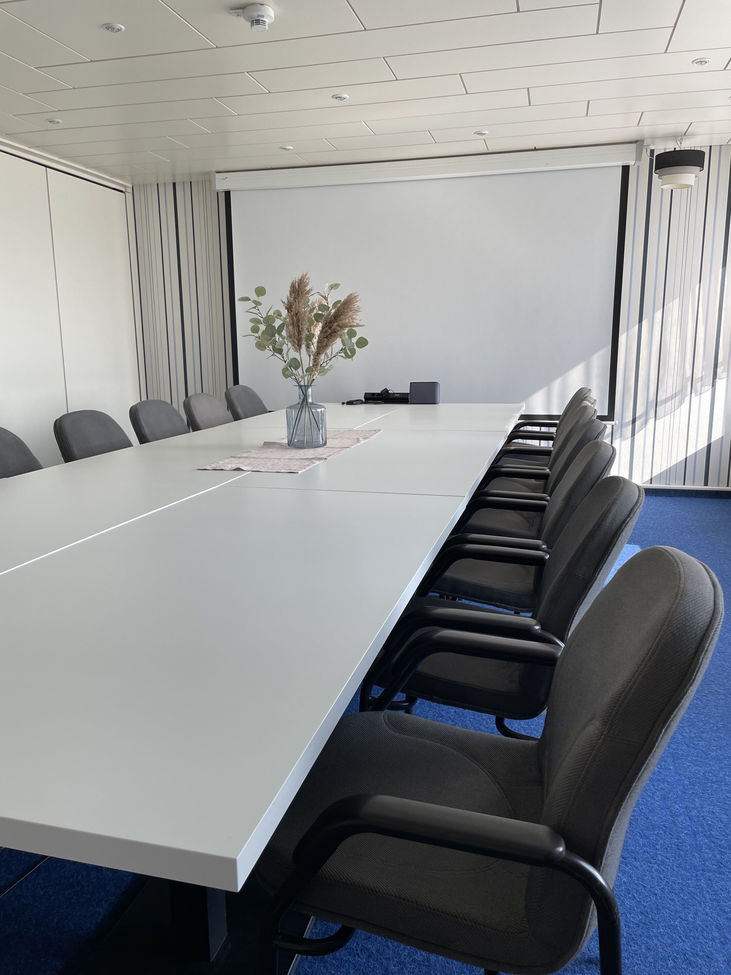 Uma sala de conferências com uma mesa comprida, várias cadeiras, uma tela de projetor e um vaso com plantas secas.