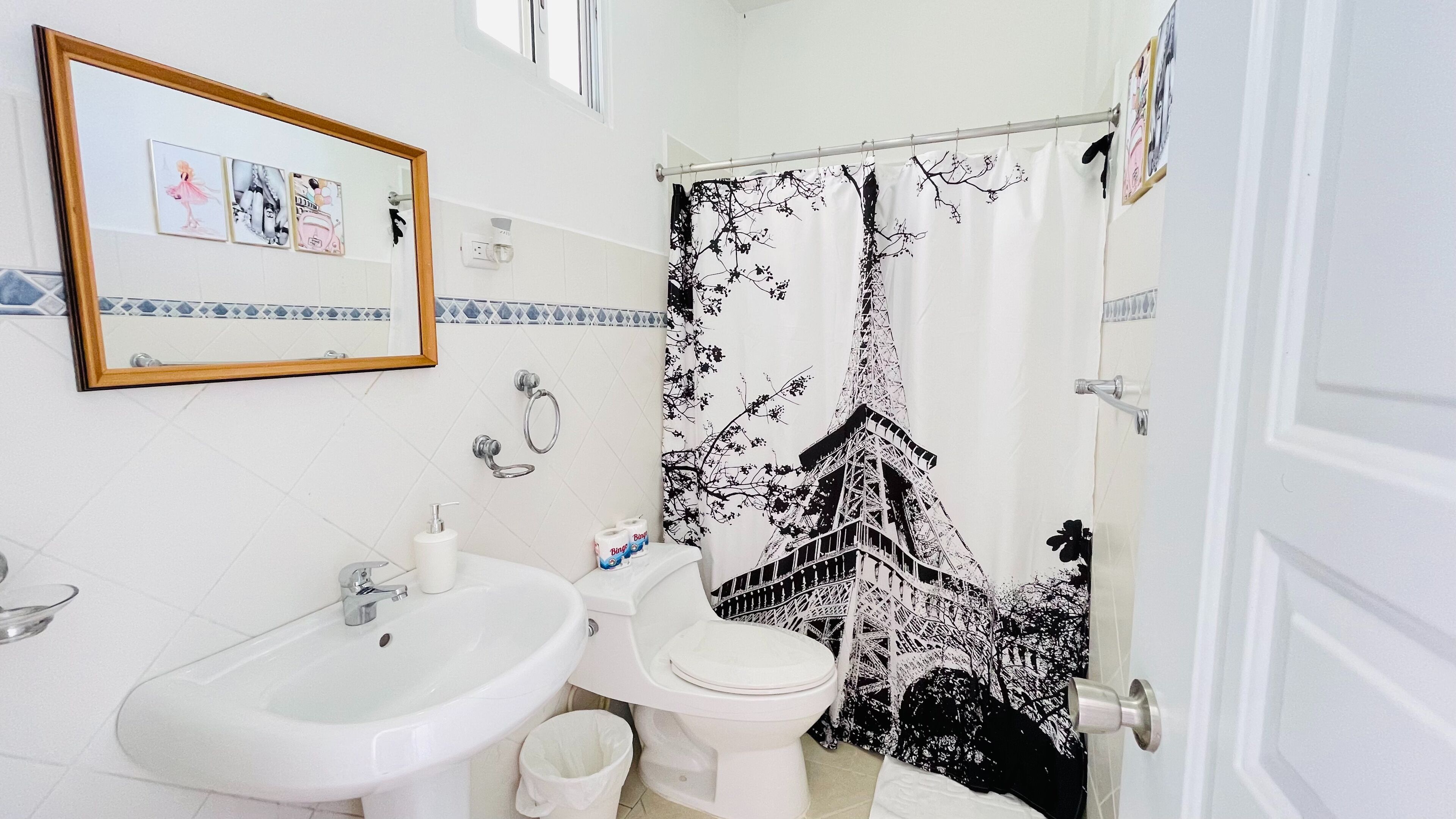 Casa de banho com lavatório branco, sanita e cortina de chuveiro com padrão de árvore. Existem quadros nas paredes e um espelho sobre o lavatório.