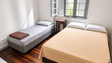 Quarto com duas camas, uma janela com persianas, uma mesa pequena e soalho de madeira.