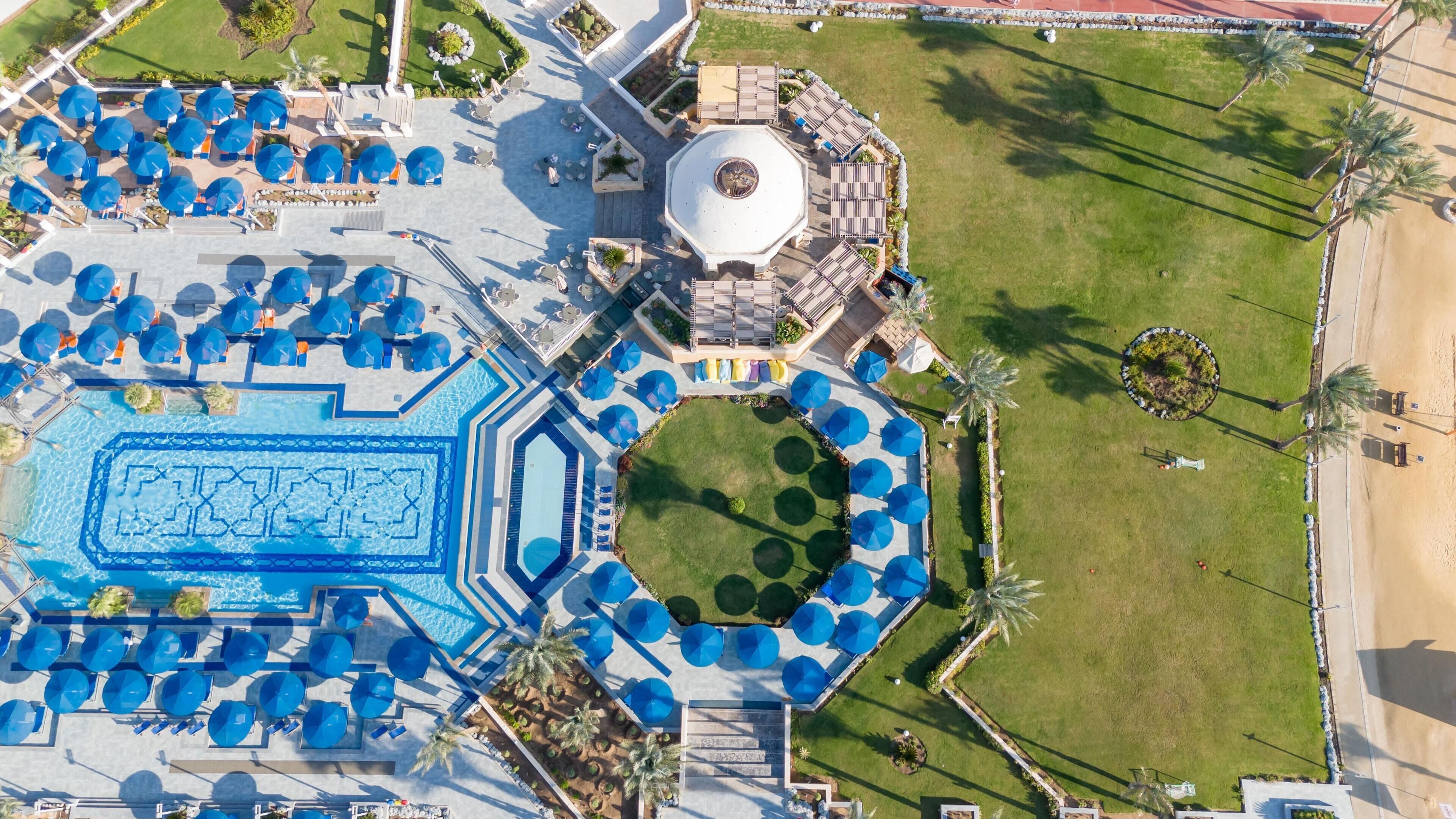 Vista aérea de um resort com piscina central, várias áreas de estar com guarda-sóis e um jardim circular.