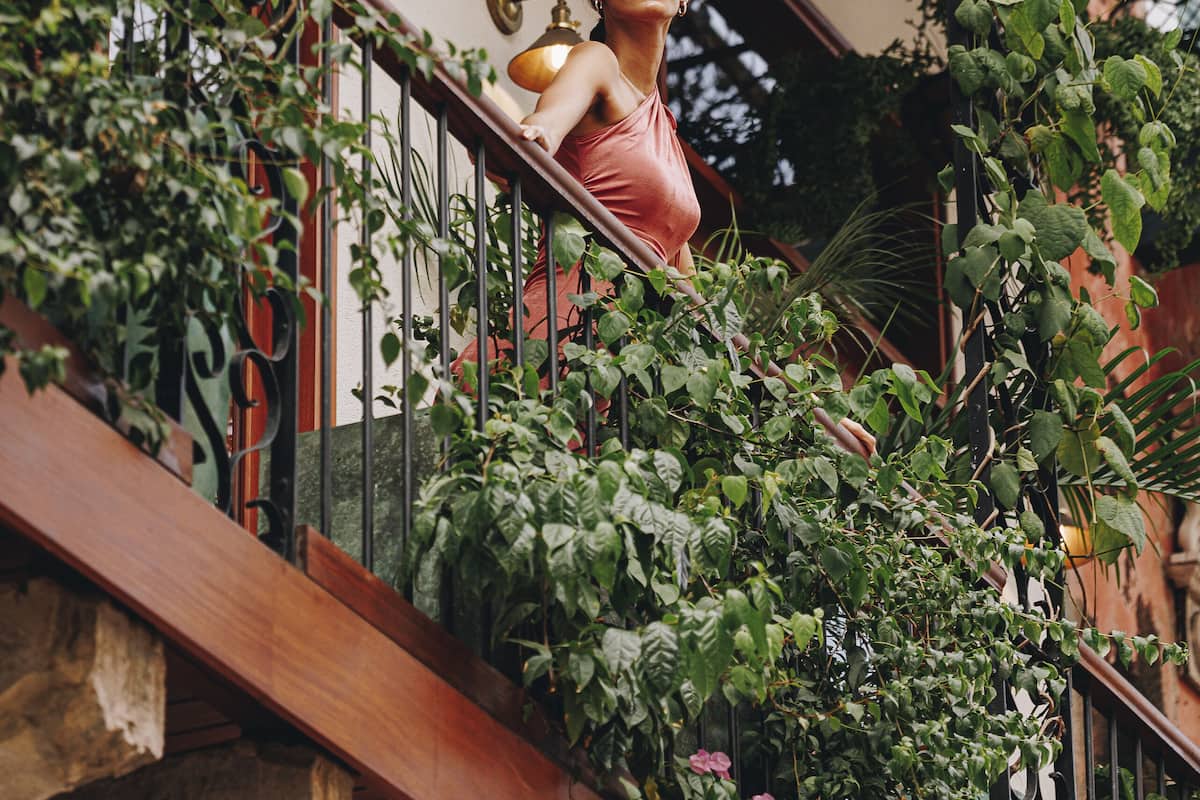 Uma varanda com vegetação exuberante, um corrimão de madeira e uma pessoa vestida de rosa.