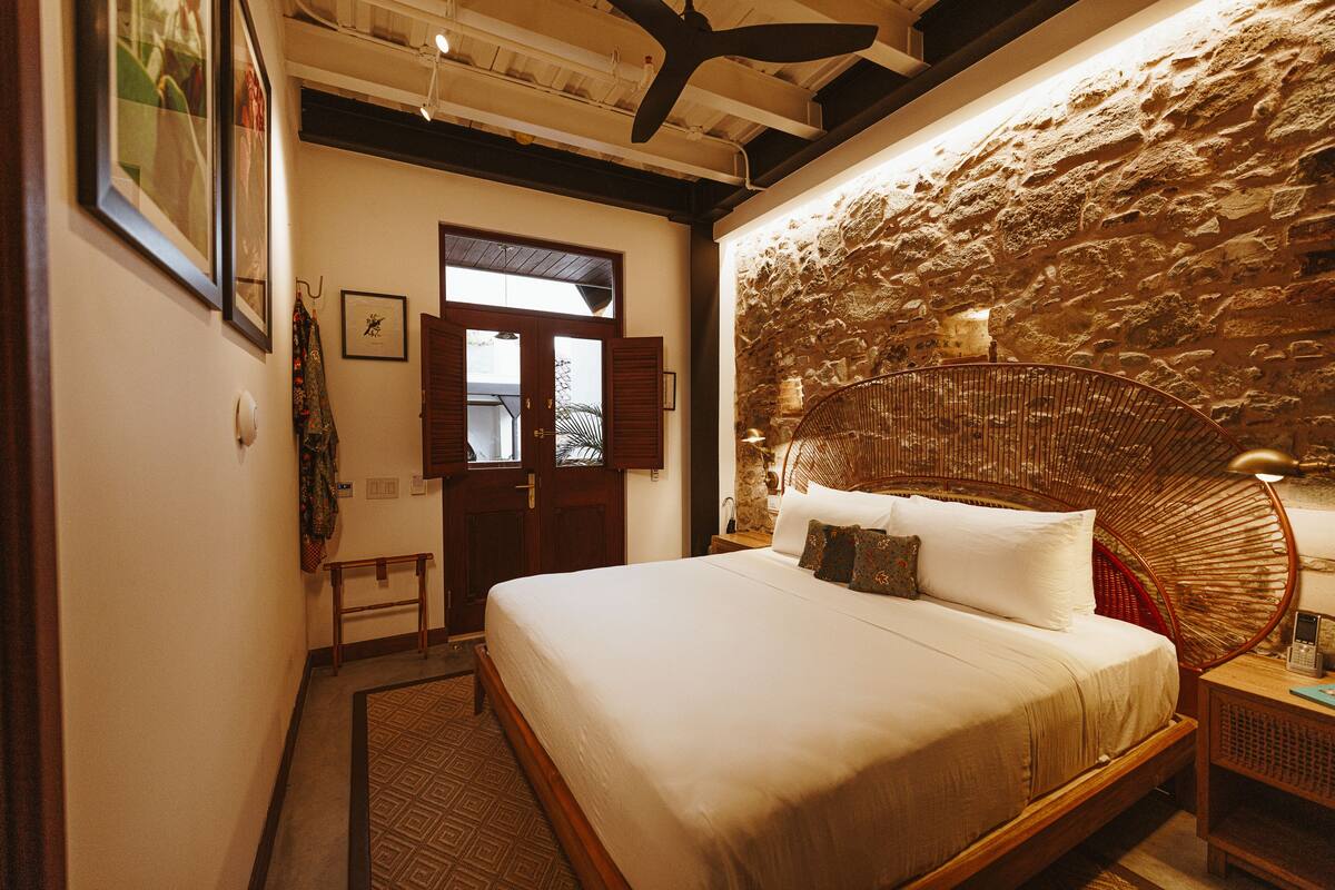 Quarto com uma cama grande, parede de pedra, ventoinha de teto de madeira e uma porta com portadas.