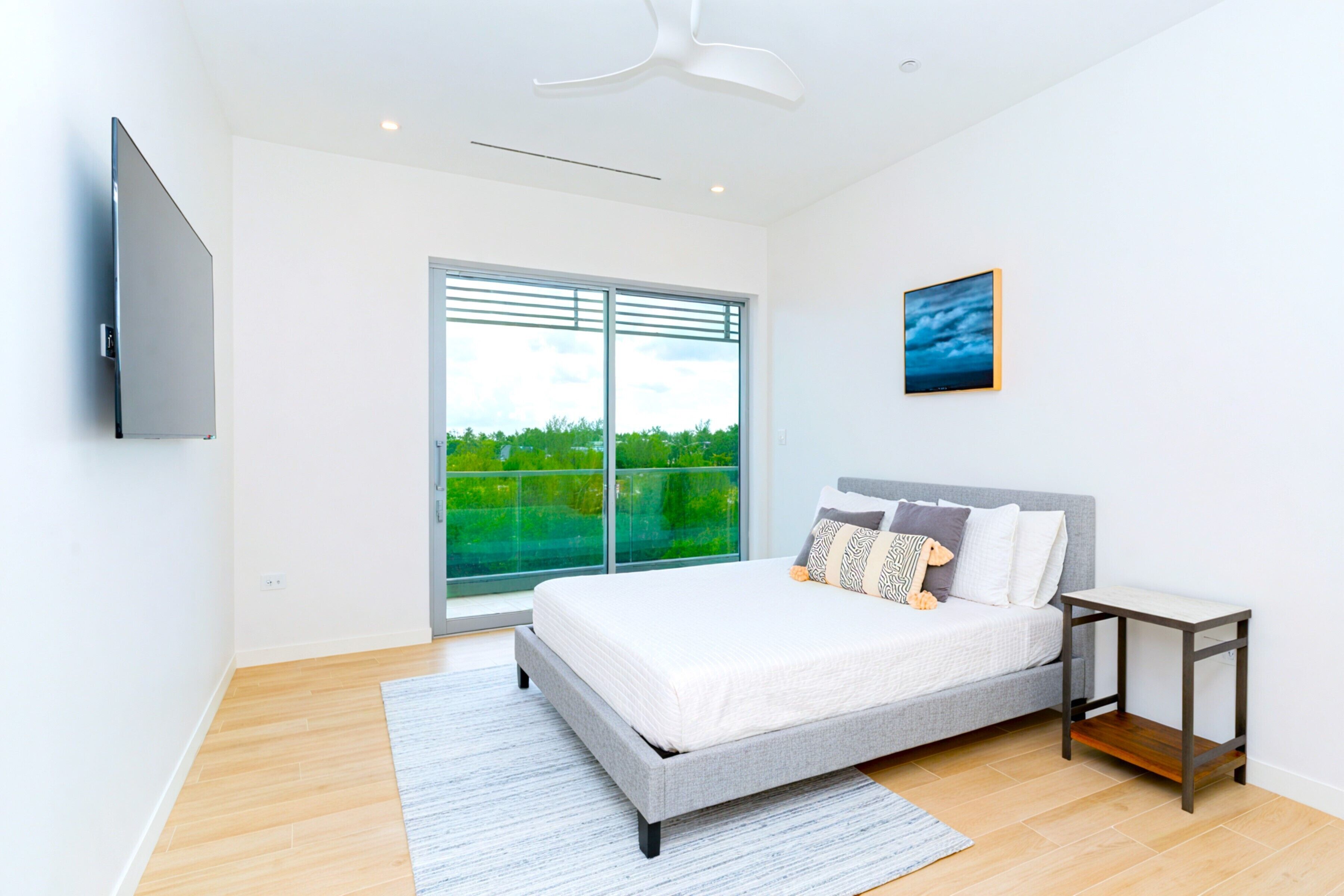 Quarto moderno com cama, mesa de cabeceira, TV na parede e uma grande janela com vista para o verde.