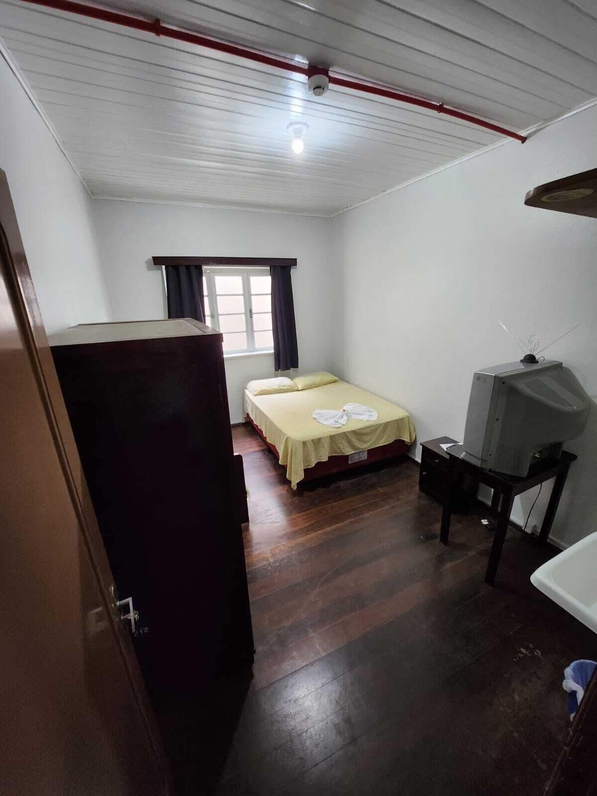 Quarto pequeno e simples, com uma cama, televisão sobre um suporte e uma janela com cortinas.