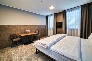 Quarto de hotel com cama, escrivaninha, duas cadeiras, uma mesa pequena com um vaso de flores e televisão na parede.