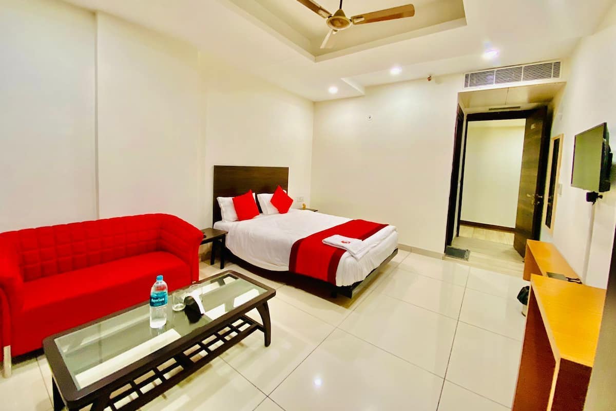 Quarto de hotel com cama, sofá vermelho, mesa de centro de vidro, ventoinha no teto e televisão na parede.