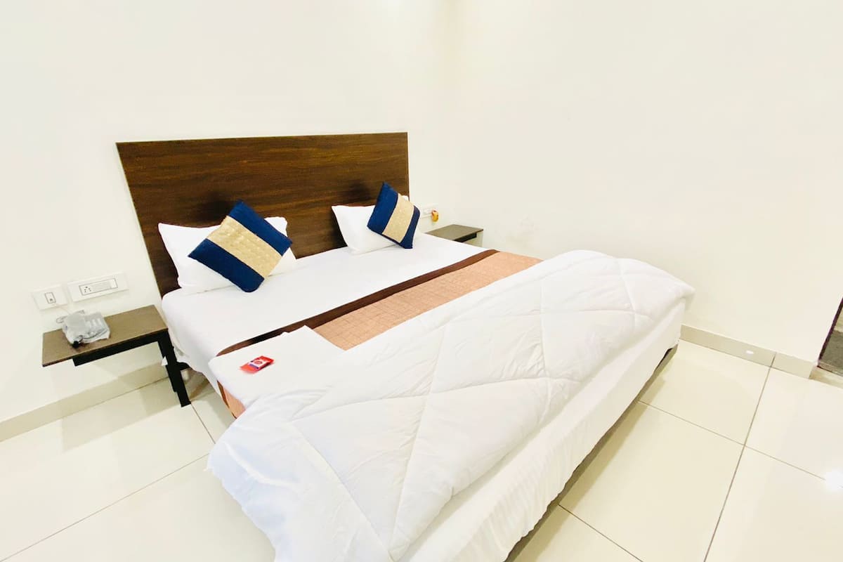 Cama bem arrumada com roupa de cama branca e almofadas azuis e bege, cabeceira de madeira, mesa de cabeceira com abajur e tomada na parede.