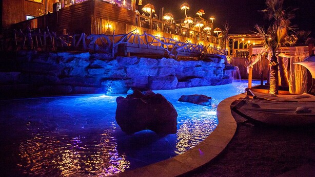 Vista noturna de um resort com piscina, deck de madeira e um edifício com janelas iluminadas.