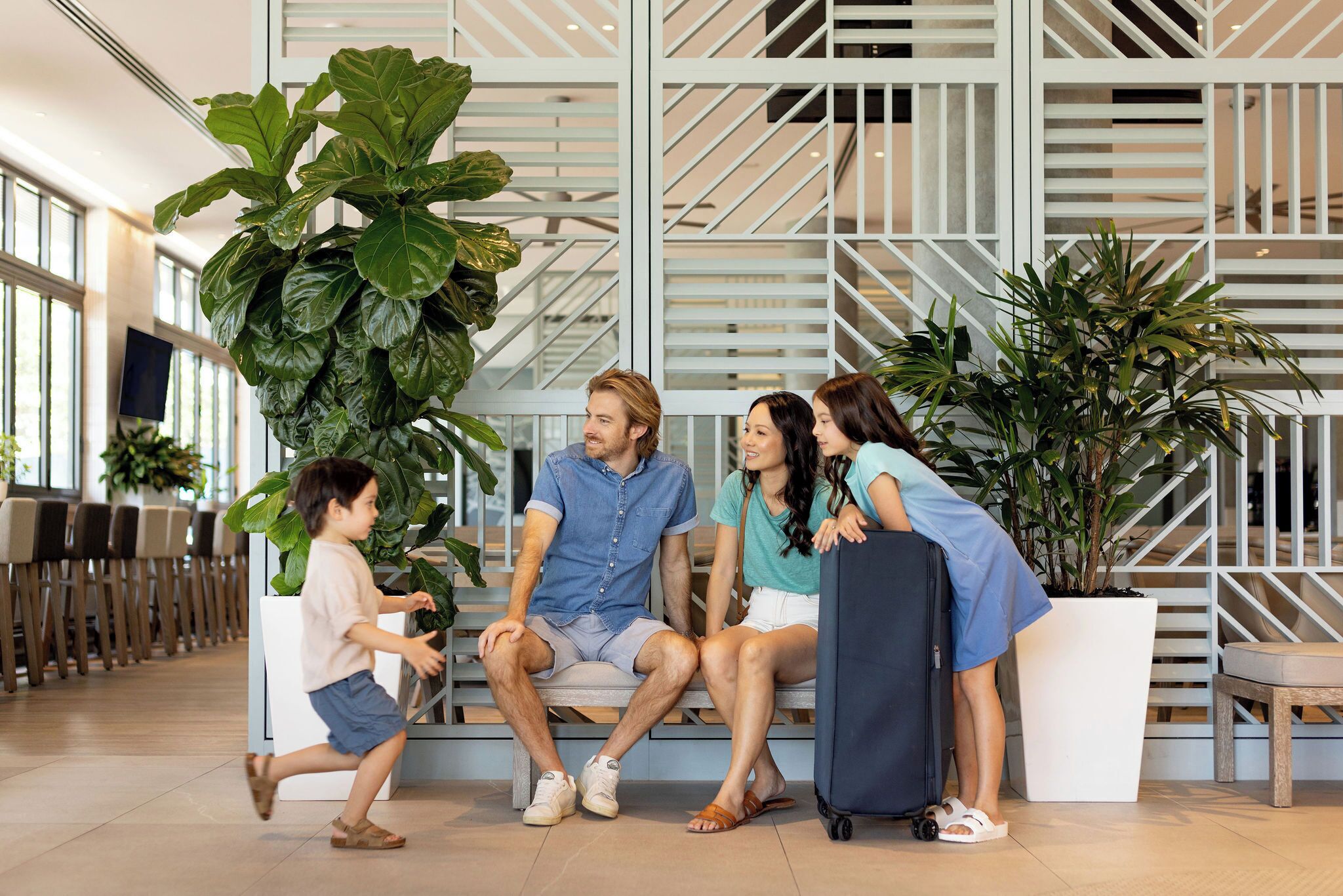 Uma família sentada num banco em espaço interior moderno, com grandes vasos de plantas e uma escadaria ao fundo.