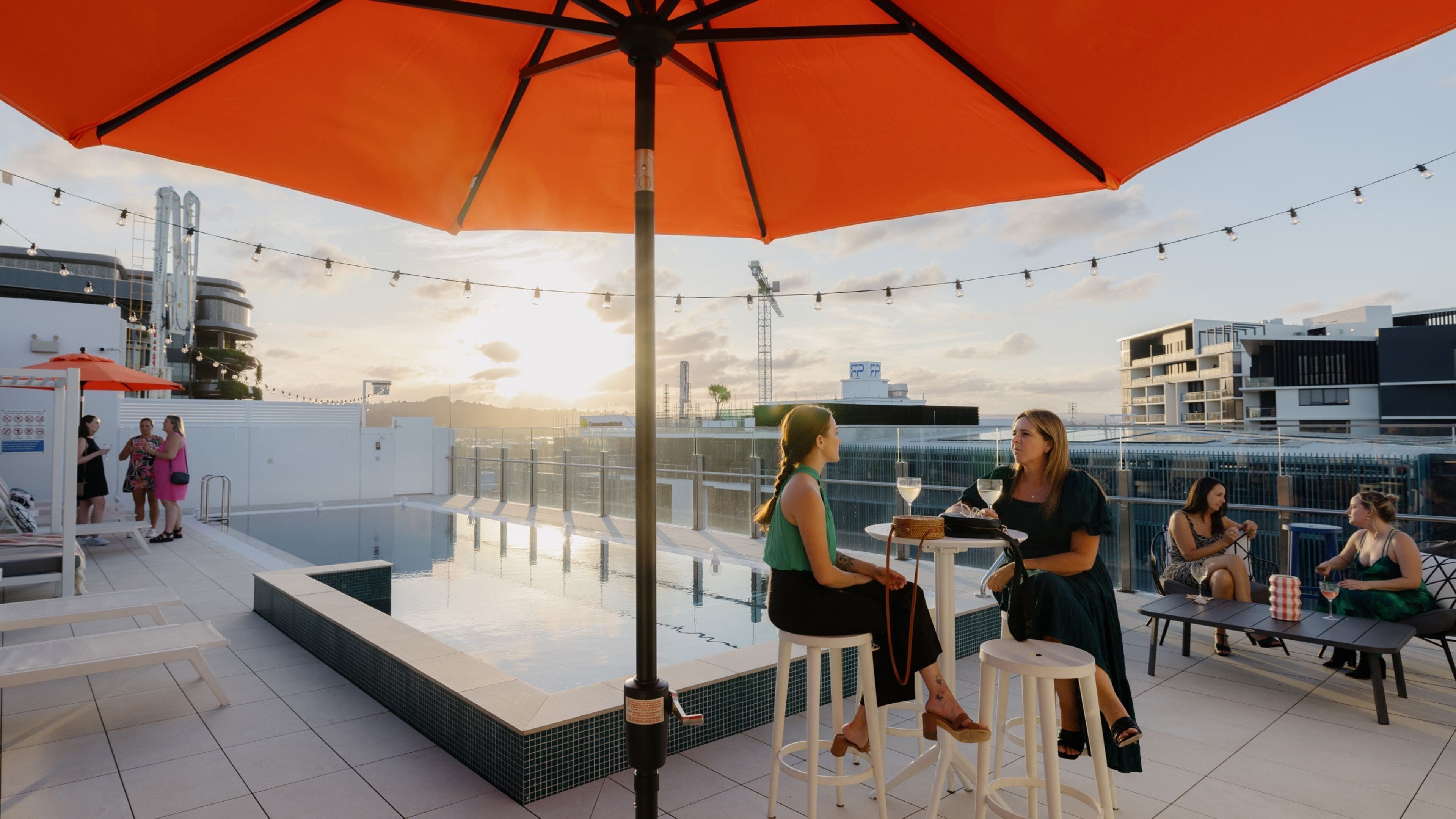 Um bar no terraço com piscina, mesas ao ar livre e um grande guarda-sol laranja.