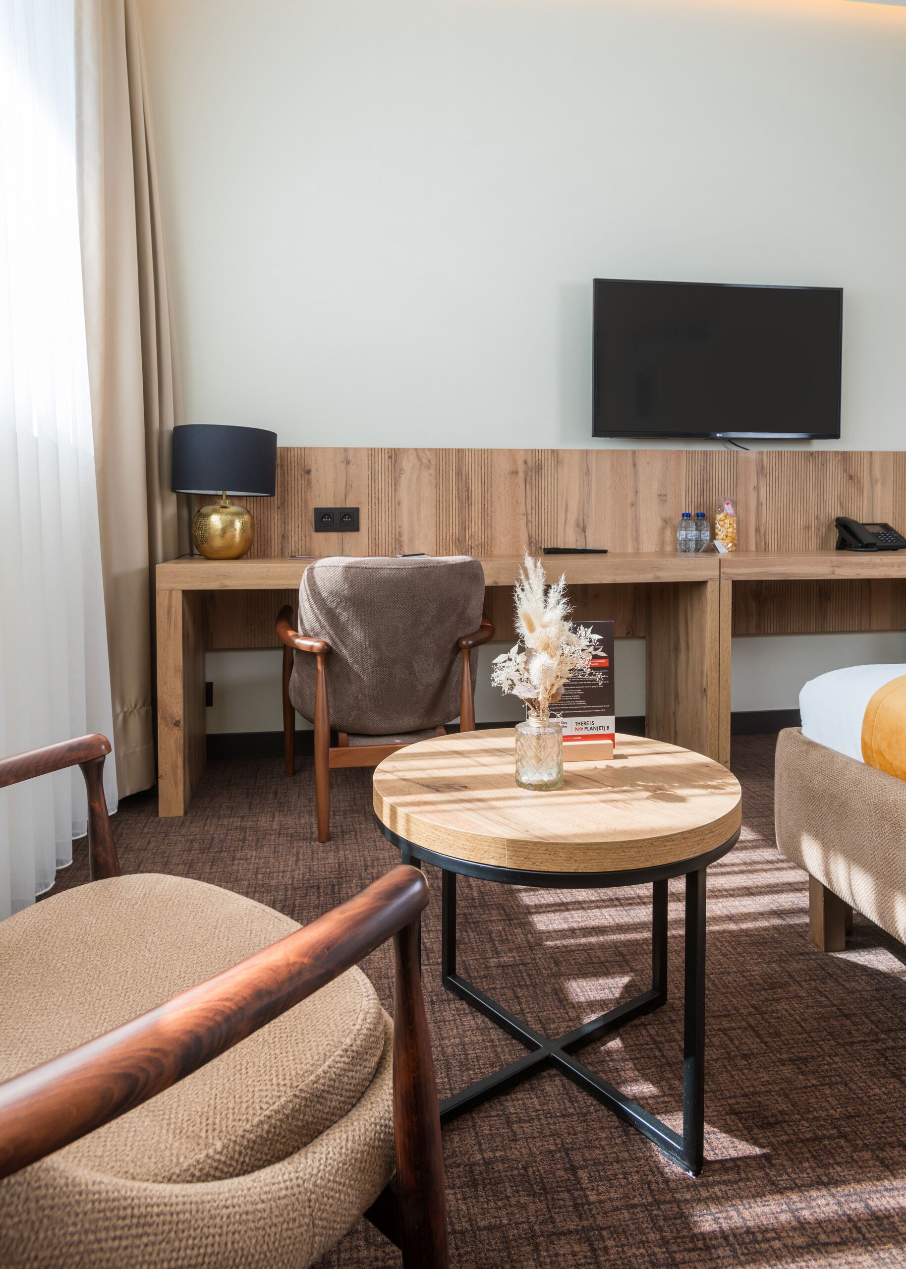 Quarto de hotel com escrivaninha de madeira, uma cadeira, uma mesa redonda com um vaso, uma cama com travesseiro amarelo e uma televisão na parede.