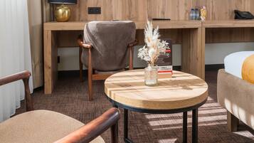 Quarto de hotel com escrivaninha de madeira, uma cadeira, uma mesa redonda com um vaso, uma cama com travesseiro amarelo e uma televisão na parede.