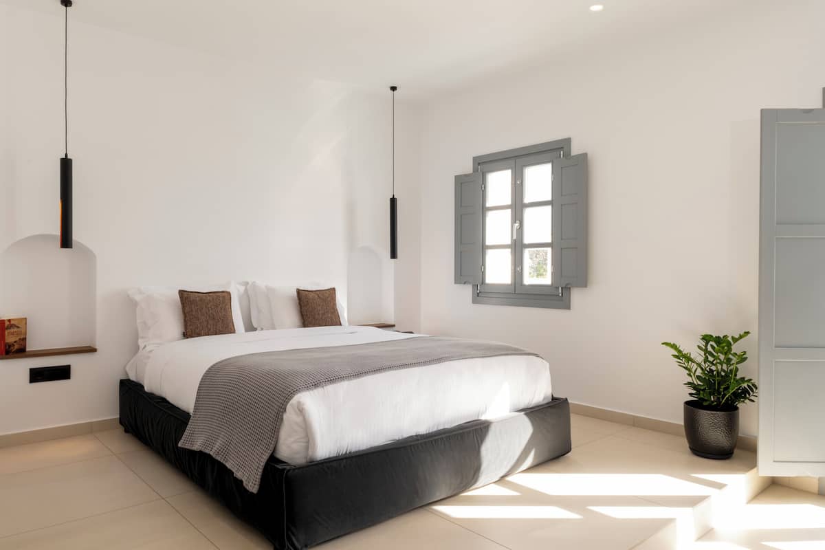 Quarto moderno com uma cama grande, luminárias pendentes, uma janela com persianas e uma planta em vaso.