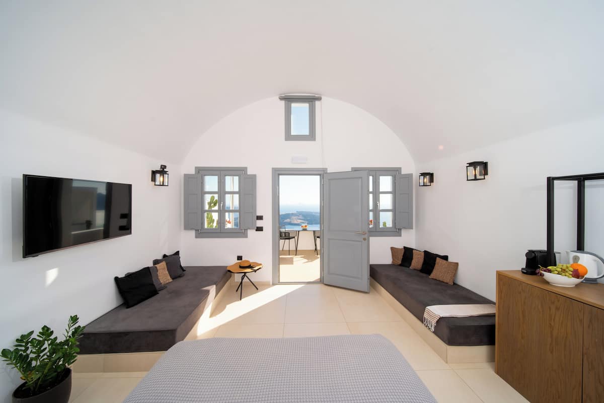 Quarto de hotel moderno com uma cama grande, televisão, sofá, mesa de jantar e vista para o mar.