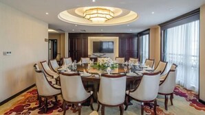 Uma sala de conferências com uma grande mesa oval, diversas cadeiras, um candelabro, uma TV de tela plana e janelas amplas com cortinas.