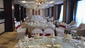 Salão de banquetes com mesas redondas preparadas para um evento formal, com cadeiras brancas, copos e um lustre.