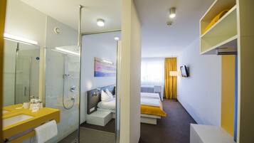 Quarto de hotel moderno com chuveiro, pia, cama, televisão e mesa de cabeceira.